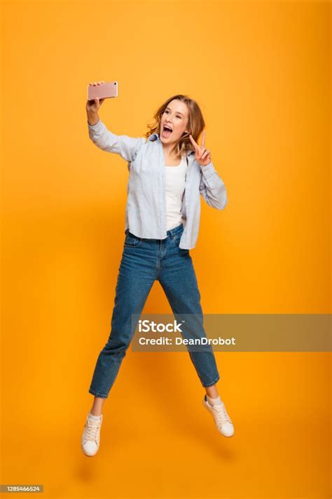 노란 배경에 고립 된 휴대 전화로 셀카를 찍는 동안 웃고 승리 사인을 보여주는 젊은 매력적인 여성의 전체 길이 초상화 갈색에 대한 스톡 사진 및 기타 이미지 Istock