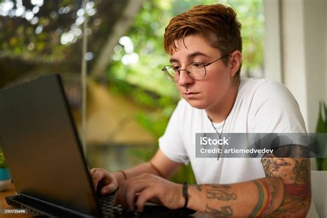 트랜스젠더 남성은 노트북을 사용하고 안경을 쓰고 문신을 한 채 집에서 프리랜서 작업에 집중합니다 원격 근무 환경의 성별 다양성 재택 식물이 있는 포용적인 직장 분위기 가상