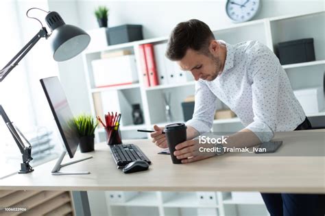 젊은 남자가 사무실에 테이블 근처 연필와 커피 한 잔을 들고 서 있다 젊은 사람이 문서와 컴퓨터와 함께 작동합니다 감독에 대한 스톡 사진 및 기타 이미지 Istock