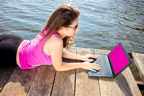 Молодая женщина нося в розовой рубашке сидя на мосте озера используя портативный компьютер