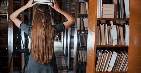 쾌활한 소녀는 그녀의 머리에 책을 넣는 동안 재미를 느낍니다 여학생이 도서관에 있습니다 교육의 개념 사진