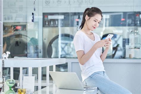 셀프 서비스 세탁실에서 세탁기와 건조기를 배경으로 노트북 과 핸드폰을 보며 일 또는 동영상 시청하는 젊은 아시아 한국 여자 모델 Stock 写真 Adobe Stock