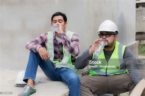 안전 반사 조끼와 헬멧을 착용 한 두 명의 아시아 건설 노동자가 피곤하고 목이 마르고 플라스틱 병의 수분을 마시는 식수가 과열된 화창한 날 건설 현장에 앉아 휴식을 취하고