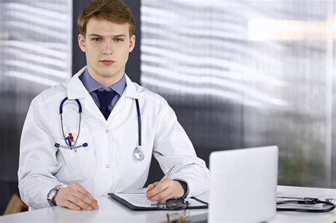 젊은 남성 의사는 앉아서 그의 직장에서 병원에서 약물 역사 기록의 클립 보드와 함께 작업 직장에서 금발 의사 완벽한 의료 서비스 의학 개념 18 19세에 대한 스톡 사진 및