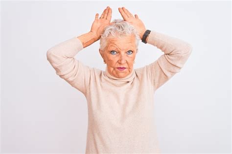 고립 된 흰색 배경 위에 서있는 터틀넥 스웨터를 입은 선임 회색 머리 여성은 손 손바닥으로 냉소적이고 회의적인 모습으로 토끼 귀 제스처를하고 있습니다 부활절 토끼 개념입니다