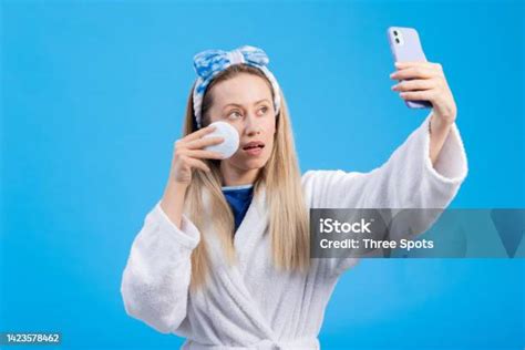 영향력있는 소녀는 아침 얼굴 관리 중에 자신의 사진을 찍고 비디오를 녹화합니다 푸른 색의 스튜디오 배경에 목욕 가운을 입은 젊은 여성 깨끗한에 대한 스톡 사진 및 기타