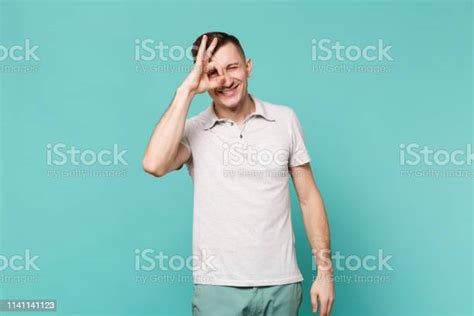 스튜디오에서 파란색 청록색 배경에 고립 된 눈 근처에 확인 제스처를 보여주는 캐주얼 한 옷에 웃는 젊은 남자의 초상화 사람들은