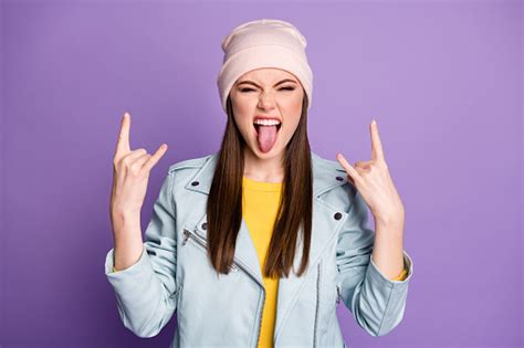 사진 의 미친 펑키 예쁜 여자 고집 혀 밖으로 입 괴롭히는 행동 로커 보여주는 뿔 손 착용 캐주얼 모자 블루 모던 재킷 고립 된 보라색 색상 배경 가죽에 대한 스톡 사진 및