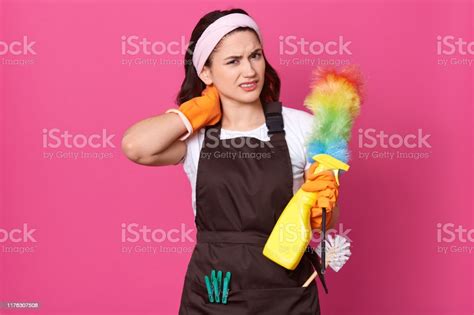 피곤 한 매력적인 젊은 백인 여성의 초상화를 닫습니다 목에 통증이 오렌지 장갑과 갈색 앞치마를 입고 서서 세제와 Pp 먼지를 들고 분홍색 배경 위에 고립 가사 집안일에 대한