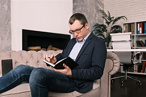 양복과 안경을 쓴 젊은 성공한 남자는 벽난로 배경에 대한 소파에 있는 넓은 사무실에 앉아 노트북이나 플래너와 함께 일합니다 성공 비즈니스 및 시간 관리의 개념 가정의 방에