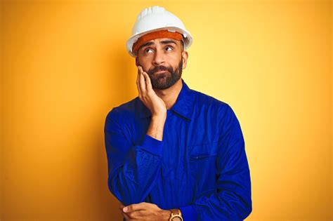 잘 생긴 인도 노동자 남자 유니폼과 헬멧 을 입고 고립 된 노란색 배경 생각 피곤 하 고 교차 된 팔우울증 문제로 지루 해 보이는 생각 사색에 대한 스톡 사진 및 기타