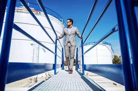 다리를 가로 질러 걷는 정장에 잘 생긴 백인 사업가의 전체 길이 배경에는 석유 저장 탱크가 있습니다 신남에 대한 스톡 사진 및