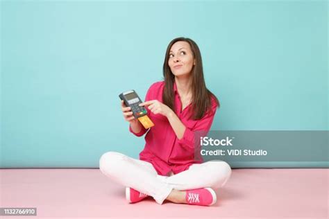 장미 셔츠에 여자의 전체 길이 초상화 흰색 바지 바닥 홀드 터미널 카드 밝은 분홍색 파란색 파스텔 벽 배경 스튜디오에 고립에 앉아 패션 라이프 스타일 개념입니다 복사 공간을