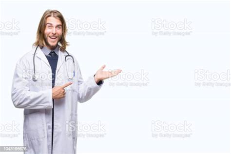 깜짝 놀라게 하 고 손으로 제시 하 고 손가락으로 가리키는 동안 카메라에 웃 고 고립 된 배경 위에 긴 머리를 가진 젊은 잘생긴 의사 남자 가리키기에 대한 스톡 사진 및