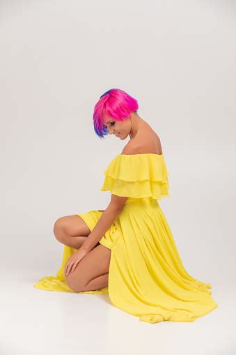 밝은 색깔의 머리를 가진 여자의 초상화 파란색 분홍색 색깔의 헤어 스타일 밝은 사이클로라마에 앉아 열린 어깨와 긴 노란색 드레스에 짧은 머리를 가진 현대 소녀 30 34세에