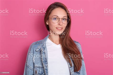 기분 좋은 찾고 여자의 실내 촬영 긍정적으로 미소 광학 안경을 입고 흰색 티셔츠와 데님 재킷 높은 첨탑에있는 분홍색 배경 위에 고립 된 포즈 모델 공간을 복사합니다 T