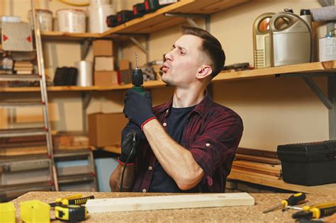 격자 무늬 셔츠 검은 티셔츠에에서 잘생긴 웃는 백인 젊은 남자 장갑 나무 나무 테이블에서 목공 워크숍에서 작업의 조각에 파워 드릴으로 드릴링 다른 도구와 장소 T 셔츠에