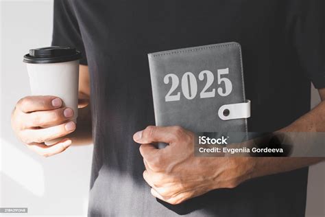 얼굴이 보이지 않는 남자 2025라는 글이 적힌 회색 일기장과 뜨거운 음료가 담긴 종이컵을 들고 있는 검은 티셔츠 배경 새해 계획 구상 계획 목표 및 계획 개념 2025년에