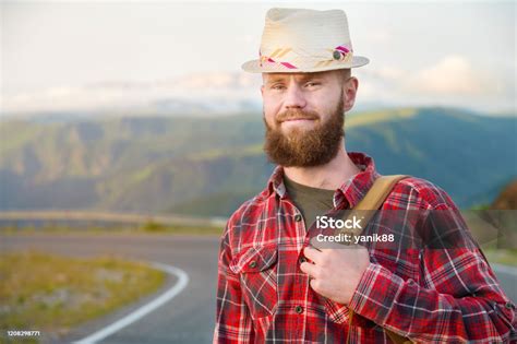 격자 무늬 셔츠를 입은 배낭과 알 수 없는 자동차 옆의 모자를 쓴 수염을 기르는 행복한 미소 여행자 힙스터의 초상화가 산의 일몰에 도로에 서 있습니다 행복하고 자신감 있는