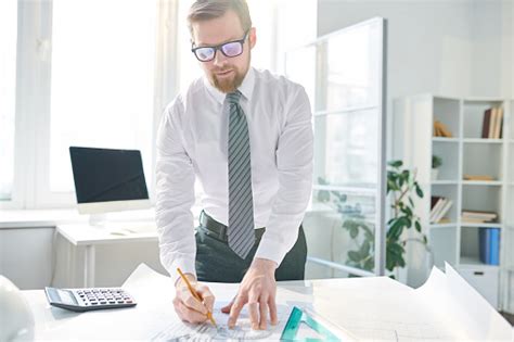 콘텐츠 바쁜 잘생긴 젊은 수염 엔지니어 책상에 서 있는 안경 및 기술 스케치를 만들기 위해 통치자를 사용 하 여 그는 사무실에서 작업 건설 산업에 대한 스톡 사진 및 기타