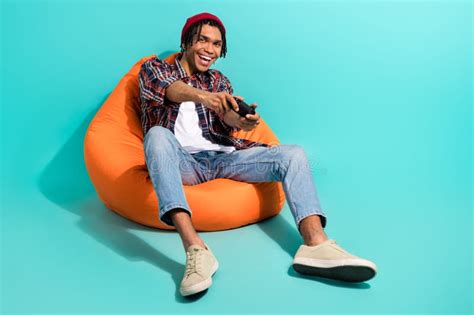 Full Length Photo Of Excited Satisfied Guy Wear Hat Sit On Bean Bag Hold Joystick Play Video