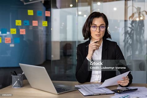 문서를 들고 안경을 쓰고 전략 계획을 세우고 디지털 도구와 다채로운 메모로 둘러싸여 비판적으로 생각하는 현대적인 사무실에서 자신감 넘치는 사업가 개념에 대한 스톡 사진 및