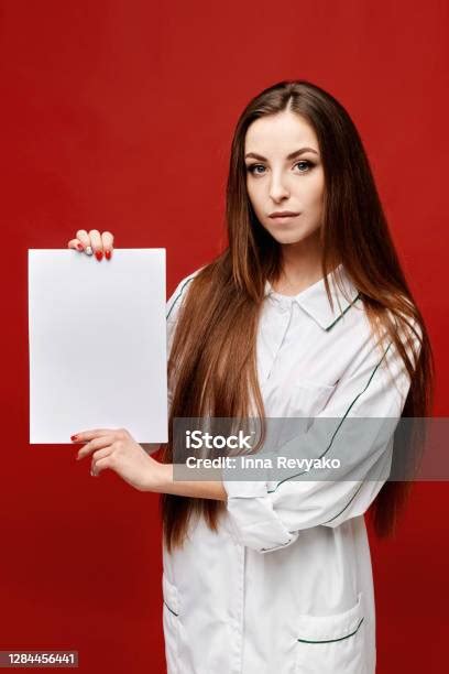 흰색 실험실 코트를 입은 젊은 여성 의사는 빨간 배경에 고립 된 그녀의 손에 빈 흰색 종이 종이를 들고 있습니다 텍스트의 공간을