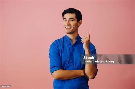 팔을 교차 한 초상화 히스패닉 라티노 남자가 복사 공간을 가리키며 검은 머리카락을 가리키며 분홍색 배경 위에 잘 생긴 젊은 성인 블루 셔츠를 입고 카메라를 바라 봅니다 30