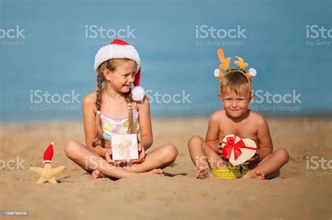 아이들은 산타 모자와 사슴 뿔을 입고 해변에 앉아있다 어린 소년과 소녀는 미소를 지으며 선물로 상자를 바라봅니다 즐거운 성탄절