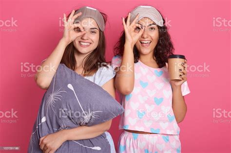 잠옷과 수면 마스크를 쓰고 분홍색 벽에 서서 행복과 기쁨을 표현하고 커피를 들고 있는 매력적인 젊은 여성들이 괜찮은 표지판을 들고 있습니다 2명에 대한 스톡 사진 및 기타