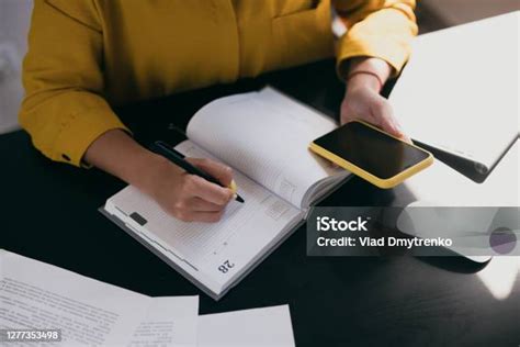 여성의 손의 시야를 잘라 노란 블라우스를 입은 백인 여성은 일기나 플래너로 글을 쓰고 전화를 보고 있다 맨 위 보기입니다 검은 책상에 종이 손에 빨간 팔찌 집에서 일하십시오