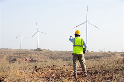 노란 헬멧을 쓰고 풍차나 풍력 터빈 농장을 가리키는 조끼를 입은 인도 엔지니어 감독관의 뒷모습 깨끗하고 녹색 에너지 전기를