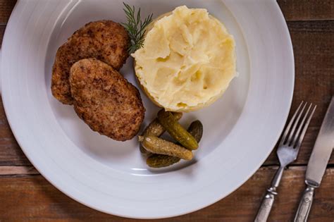 으깬 감자를 곁들인 두 개의 돼지 고기 커틀릿 딜과 피클의 장식 음식은 둥근 흰색 접시에 있습니다 접시는 나무 배경에 서 있고 포크와 나이프가 근처에 있습니다 0명에 대한