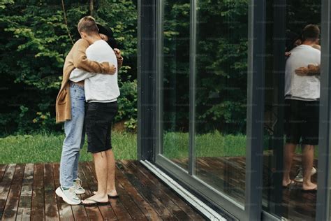 세련된 힙스터 커플은 나무 테라스에서 포옹하고 키스하며 산의 오두막에서 휴식을 취합니다 큰 창문과 나무를 배경으로 현대적인 의상을 입은 행복한 젊은 가족 사진