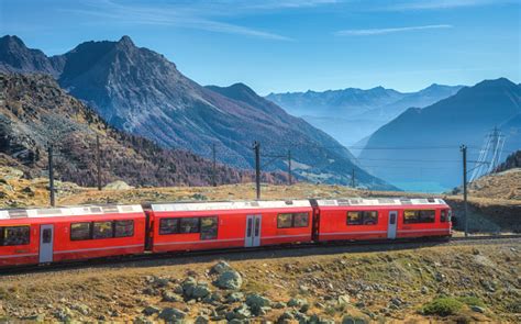 화창한 가을날 스위스 알프스를 가로지르는 빨간색 현대식 기차가 있습니다 움직이는 기차 고산 산 계곡 오렌지 언덕 가을에 일몰에 푸른 하늘이 있는 풍경 Bernina