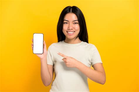 캐주얼 한 티셔츠를 입은 행복한 중국 긍정적 인 소녀는 빈 흰색 화면으로 현대 스마트 폰을 보여주고 손가락을 가리키며 고립 된 오렌지색 배경에 서서 웃고 있습니다 모형 복사