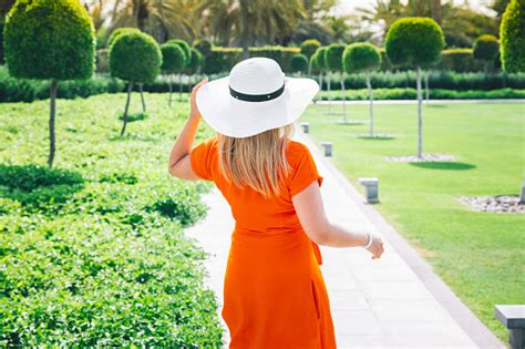 고품질 사진 빨간 드레스와 하얀 여름 모자를 쓴 젊은 백인 여성 아가씨가 자빌 공원의 길에 서서 화창한 여름날 공원의 나무에서 거리를 바라보고 있다 뒷모습 여름철 리조트 인