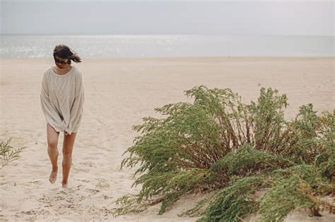 녹색 잔디와 바다의 배경에 모래 해변에서 걷는 바람이 부는 머리를 가진 아름다운 여자 조용한 조용한 순간 추운 해안에서 편안한 베이지 니트 스웨터에 세련된 젊은 여성