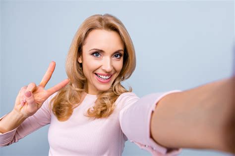 닫기 금발 곱슬 머리와 매력적인 성숙 평온한 즐거운 여자의 초상화 그녀는 Selfie를 복용 이며 회색 바탕에 고립 된 두 개의 손가락을 보여주는 가정용 캠코더에 대한 스톡