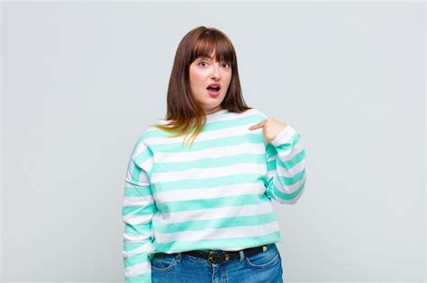 Полная женщина выглядит шокированной и удивленной с широко открытым ртом указывая на себя