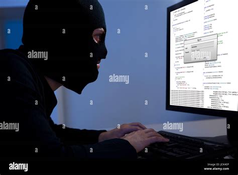 Masked Hacker Wearing A Balaclava Sitting At A Desk Downloading Private Information Off A