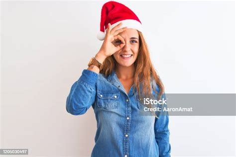 고립 된 배경 위에 크리스마스 모자를 쓰고있는 젊은 아름다운 빨간 머리 여성은 행복한 얼굴로 손가락을 통해 보는 손 미소와 함께 확인 제스처를하고 있습니다 공휴일에 대한