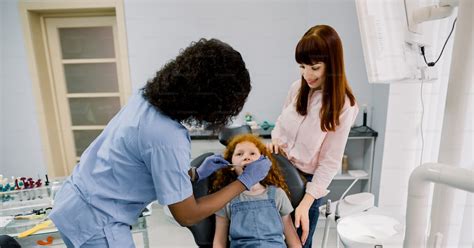 치과 도구를 가진 젊은 여성 아프리카계 미국인 치과 의사 그녀의 어린 환자 소녀의 치과 검진을 합니다 검사 중에 겁에 질린 귀여운 딸을 안심시키는 젊은 어머니 사진