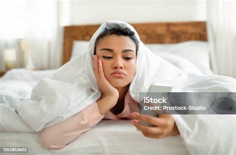 화가 젊은 흑인 여성 스마트폰 화면을 보고 하는 동안 담요 아래 침대에 누워 외로움 또는 우울 느낌 마음이 산란해진에 대한 스톡 사진 및 기타 이미지 Istock