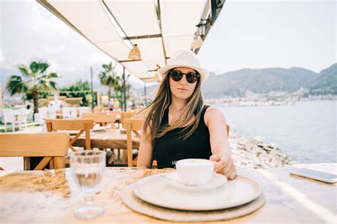 선글라스와 모자를 쓴 젊은 여성 여성이 바다와 산을 배경으로 한 거리의 야외 해변 카페 레스토랑 커피숍에서 커피를 마신다 안녕하세요 여름 휴가 휴가 거리에 대한 스톡 사진