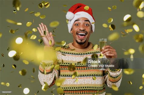 니트 스웨터 레드 크리스마스 모자에 젊은 재미 아프리카 남자는 일반 녹색 카키 배경 스튜디오에서 고립 된 색종이 반짝이에 와인 잔 음료 샴페인을 개최합니다 새해 복 많은