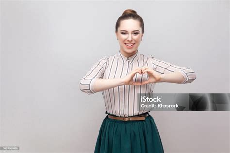 스트라이프 셔츠와 그린에서 아름 다운 젊은 여자 화장과 치마 하 고 금지 헤어스타일 마음 또는 사랑 몸짓으로 서 있는 Toothy 미소와 함께 카메라를 보고 갈색 머리에