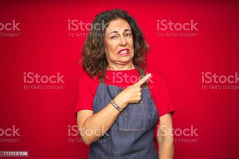 빨간 고립된 배경 위에 앞치마 유니폼을 입은 중년 시니어 여성은 집게 손가락으로 걱정하고 긴장하고 걱정하고 놀란 표정을 지으며 가리킵니다 아내에 대한 스톡 사진 및 기타