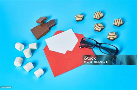 흩어진 과자 줄무늬 사탕 원통형 마시멜로 안경 근처의 깨진 초콜릿 흰색 빈 종이 시트가 있는 봉투가 파란색 책상 위에 놓여 있습니다 사랑 편지 개념입니다 클로즈업 개체