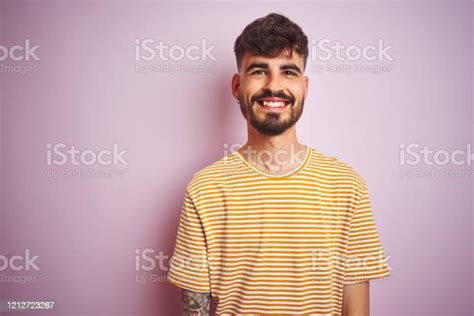 얼굴에 행복하고 시원한 미소와 고립 된 분홍색 배경 위에 서 노란색 줄무늬 티셔츠를 입고 문신을 가진 젊은 남자 운이 좋은 사람